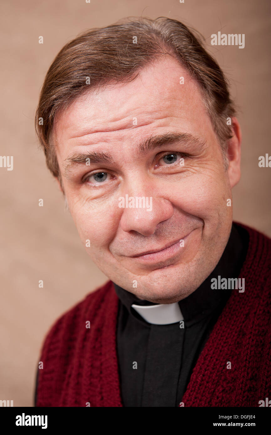 Portrait of a smiling vicar wearing a burgundy cardigan against a tan ...