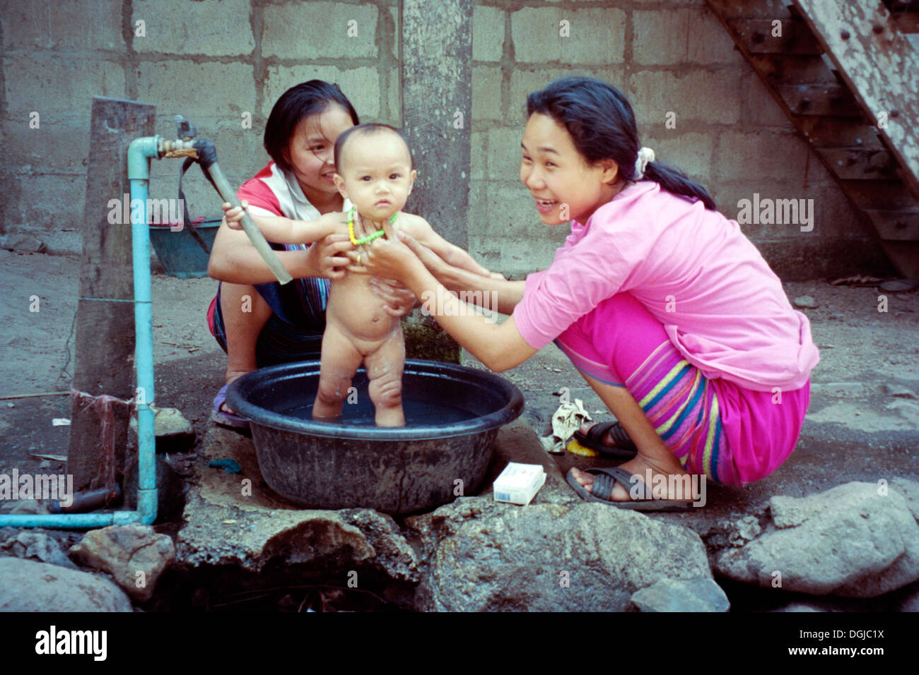two young female villagers bathing a child in a bowl using a standpipe ...