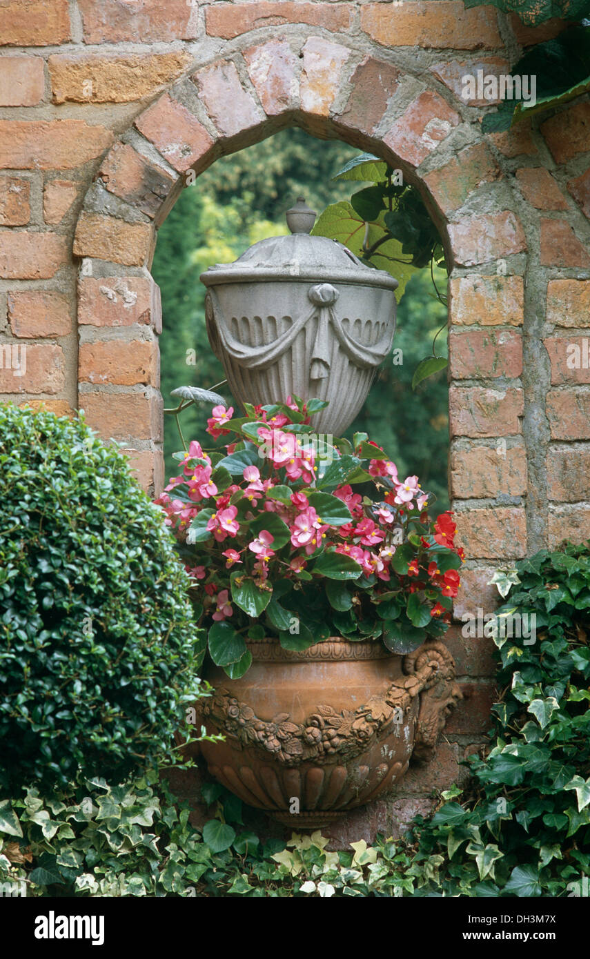Close-up of red impatiens in integral terracotta pot below stone urn in ...