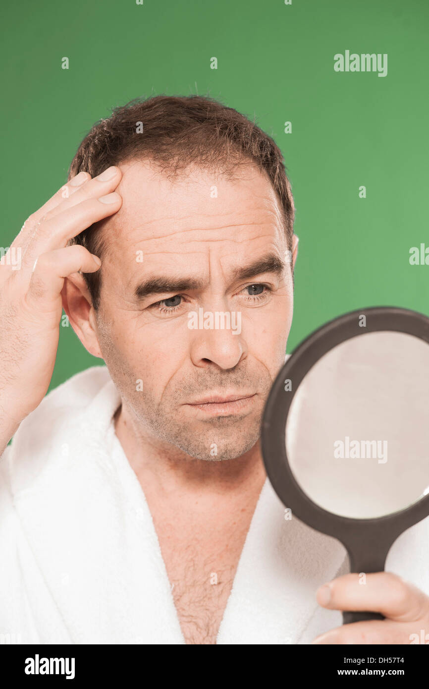 Man wearing a bathrobe observing himself critically in a hand mirror ...