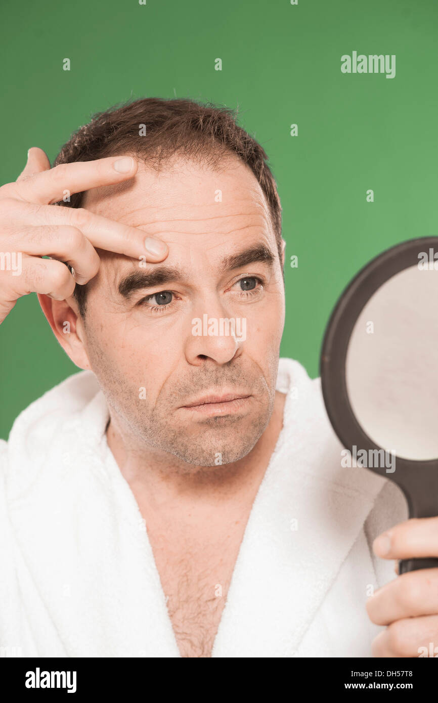 Man wearing a bathrobe observing himself critically in a hand mirror ...