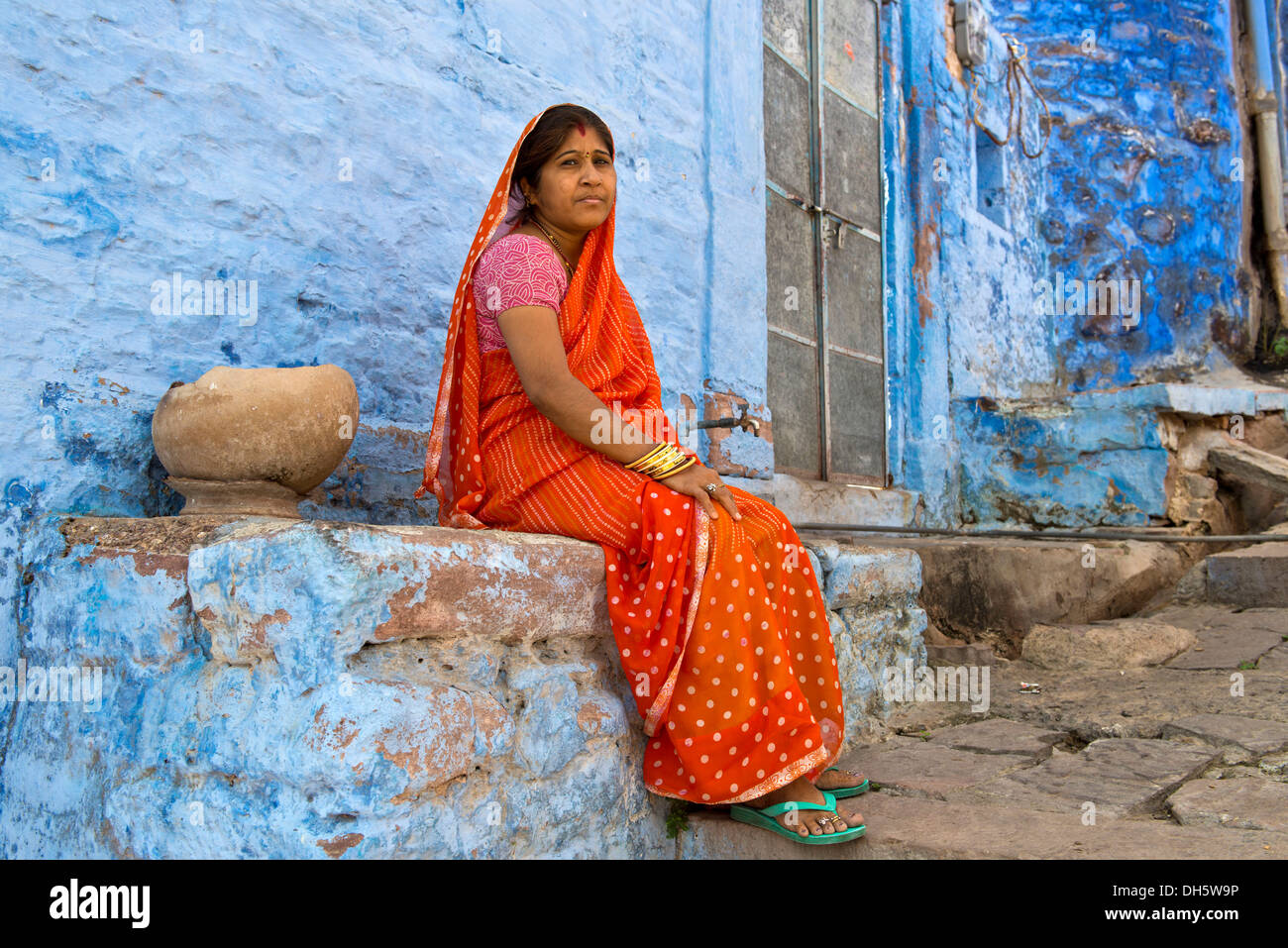 Elderly woman wearing a red sari sitting on a ledge of a blue painted ...