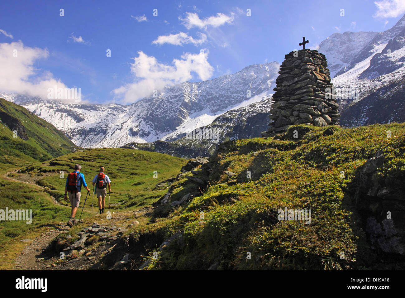Two mountain walkers walking on path along cross on stone socle in the ...