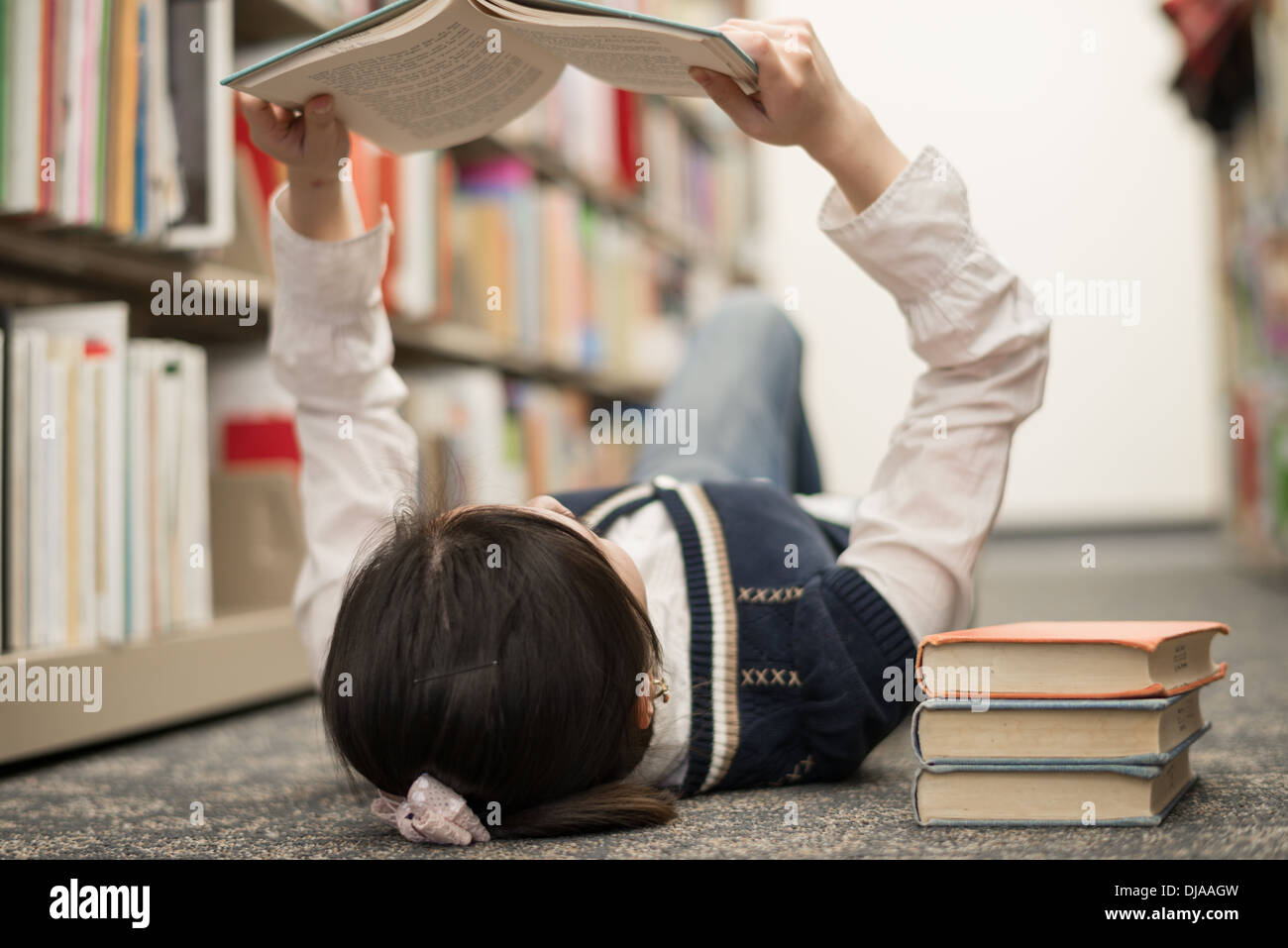 Young girl student laying on the library floor and reading a stack of ...