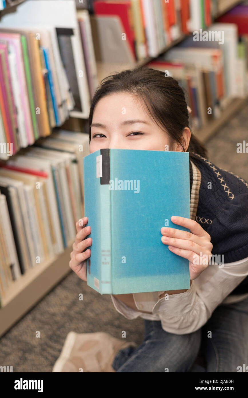 Playful student covering mouth with a blue hard cover book in library ...