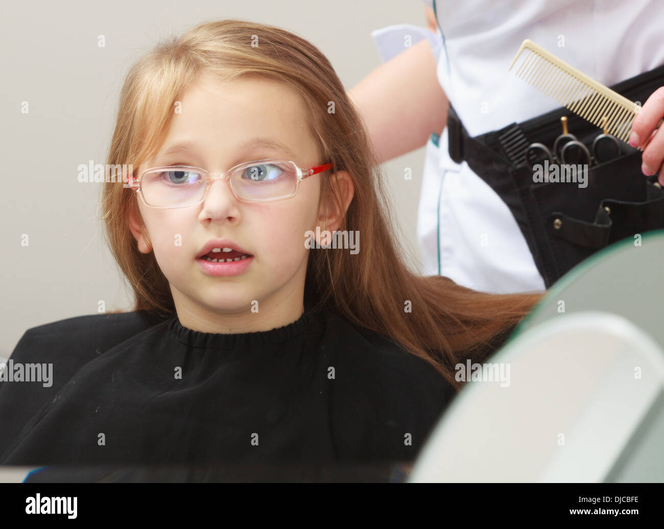 Kid by hairstylist. Hairdresser with comb combing hair of little girl in eyeglasses. Child in ...