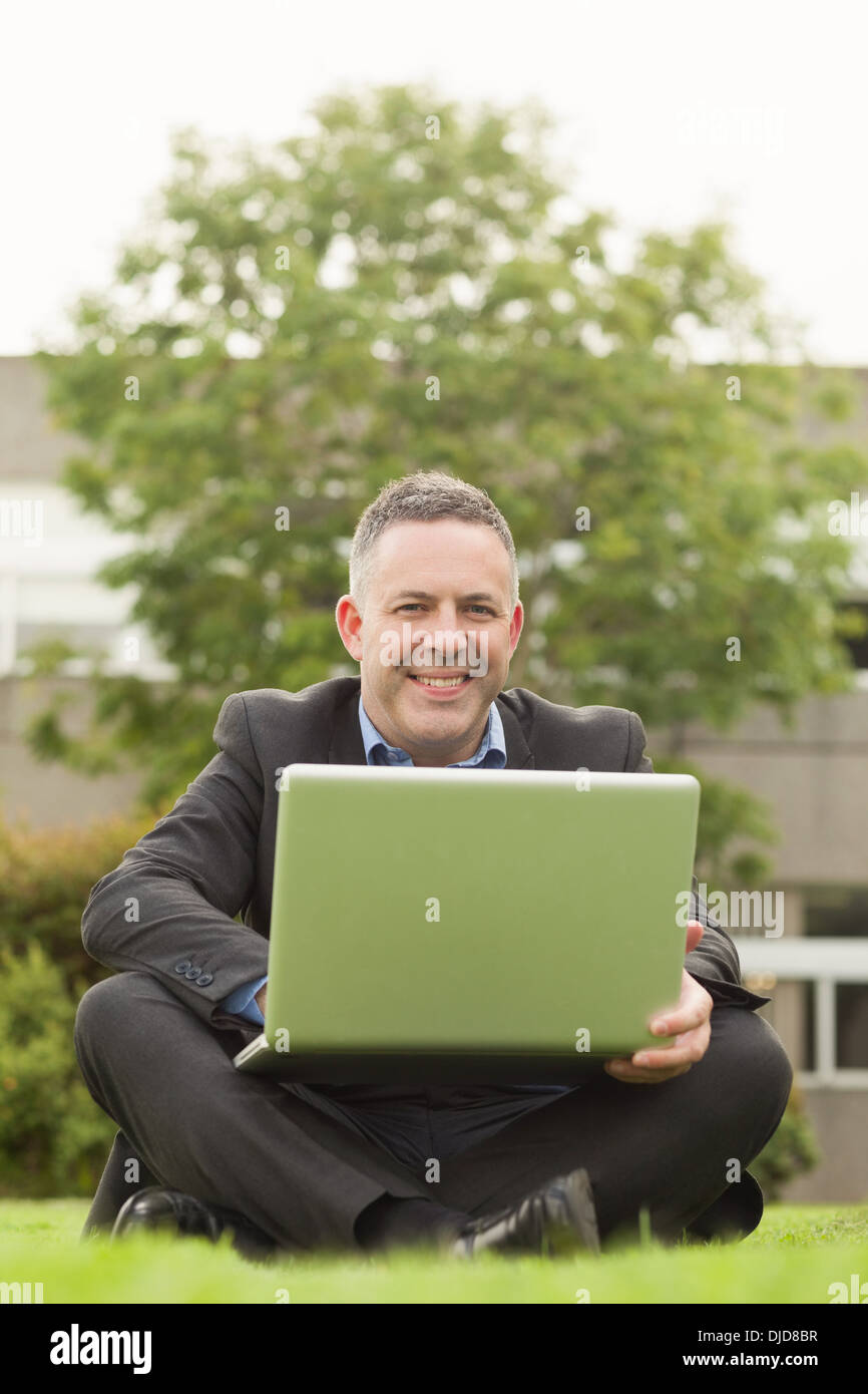 Happy professor sitting outside on campus using his laptop looking at ...
