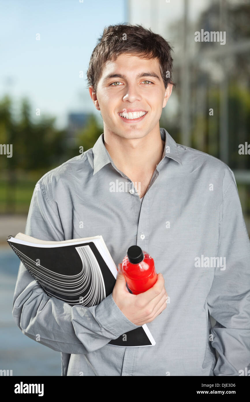 Male Student With Juice Bottle And Book Standing On Campus Stock Photo ...