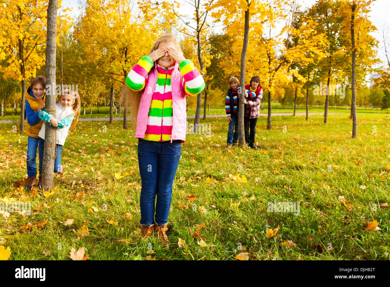 Group of five kids play hide and seek with girl counting and friends hiding behind the trees ...