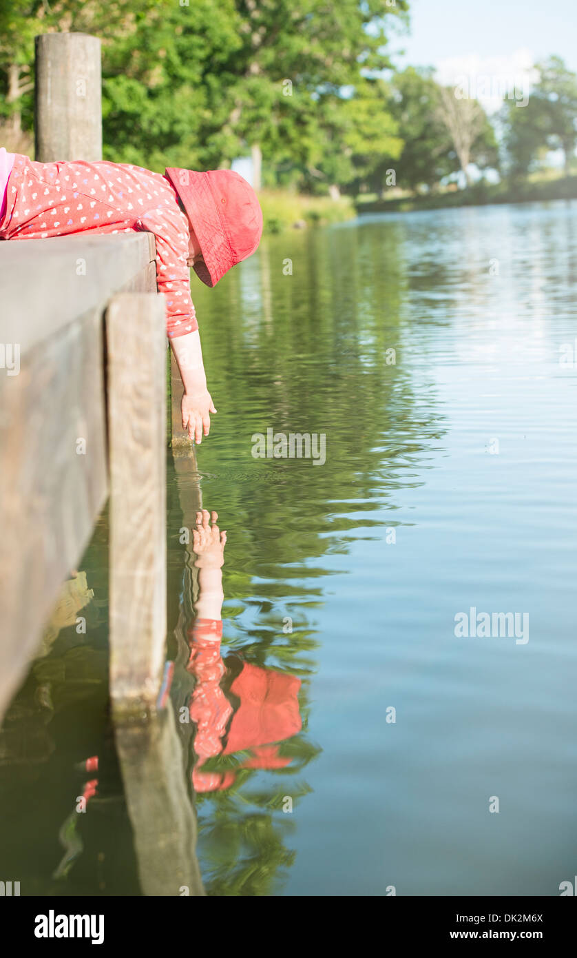 Little girl lying on jetty, looking down at the water and reaching for ...