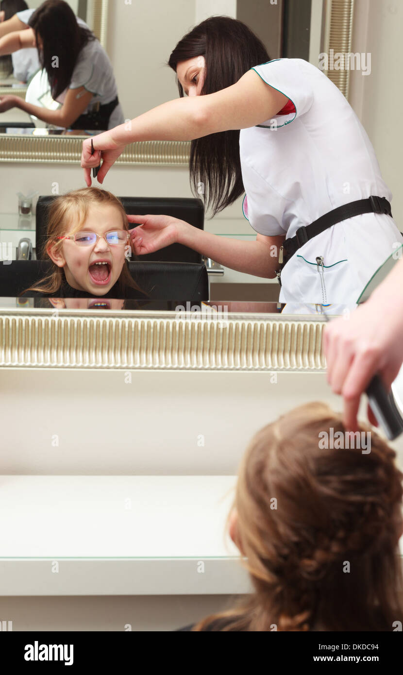 Kid by hairstylist. Hairdresser with comb combing hair of little girl in eyeglasses. Child in ...