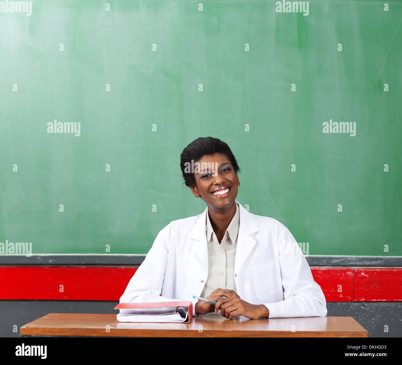 Happy Female Teacher Sitting At Desk In Classroom Stock Photo - Alamy