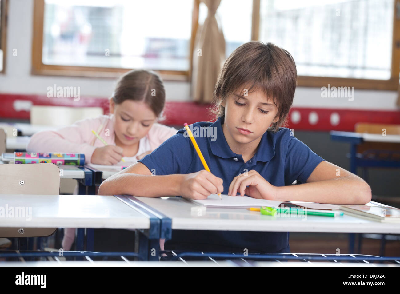 Little Boy Writing Notes With Classmate In Background Stock Photo - Alamy