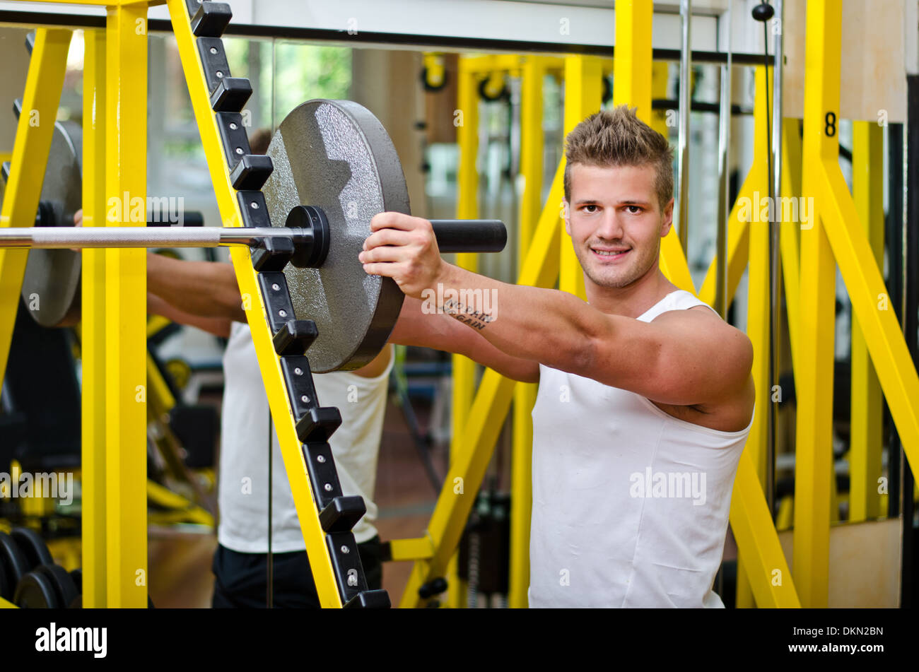 Smiling handsome young man in gym putting weight disck on barbell ...