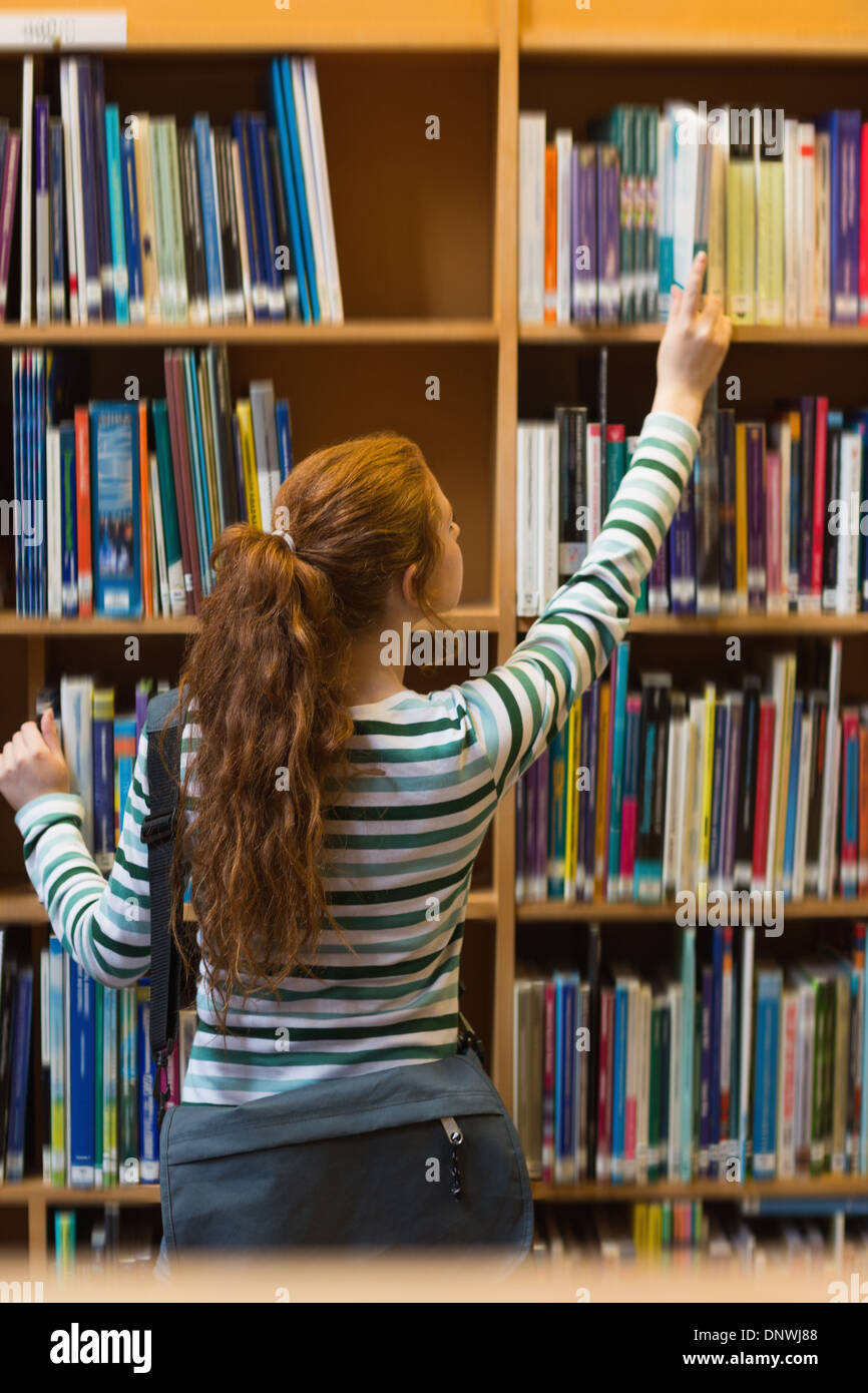 Redhead student taking book from shelf in library Stock Photo - Alamy