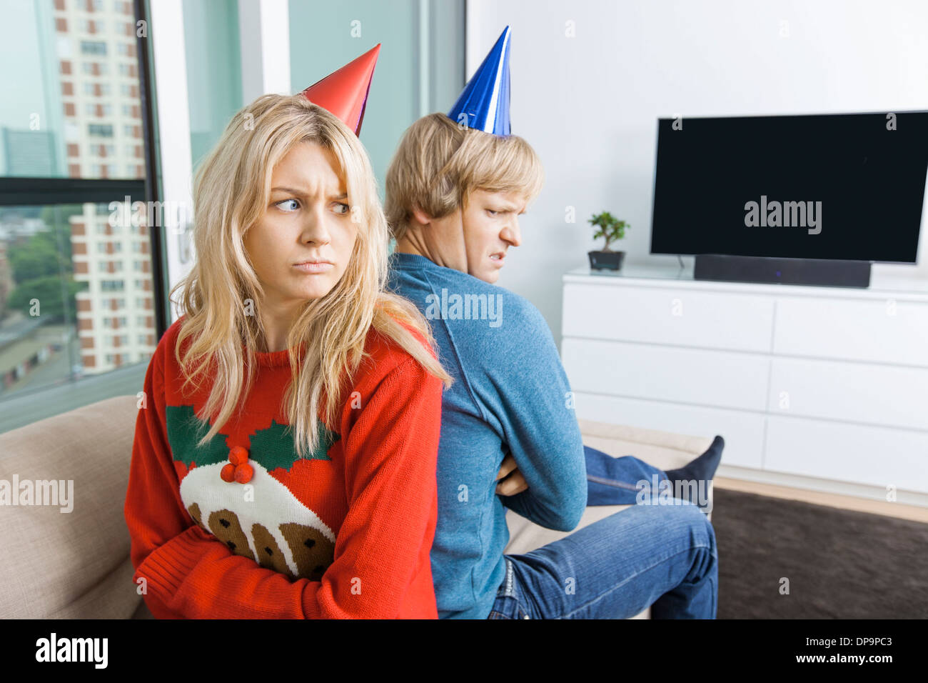 Annoyed couple in Christmas sweaters and party hats sitting back to ...