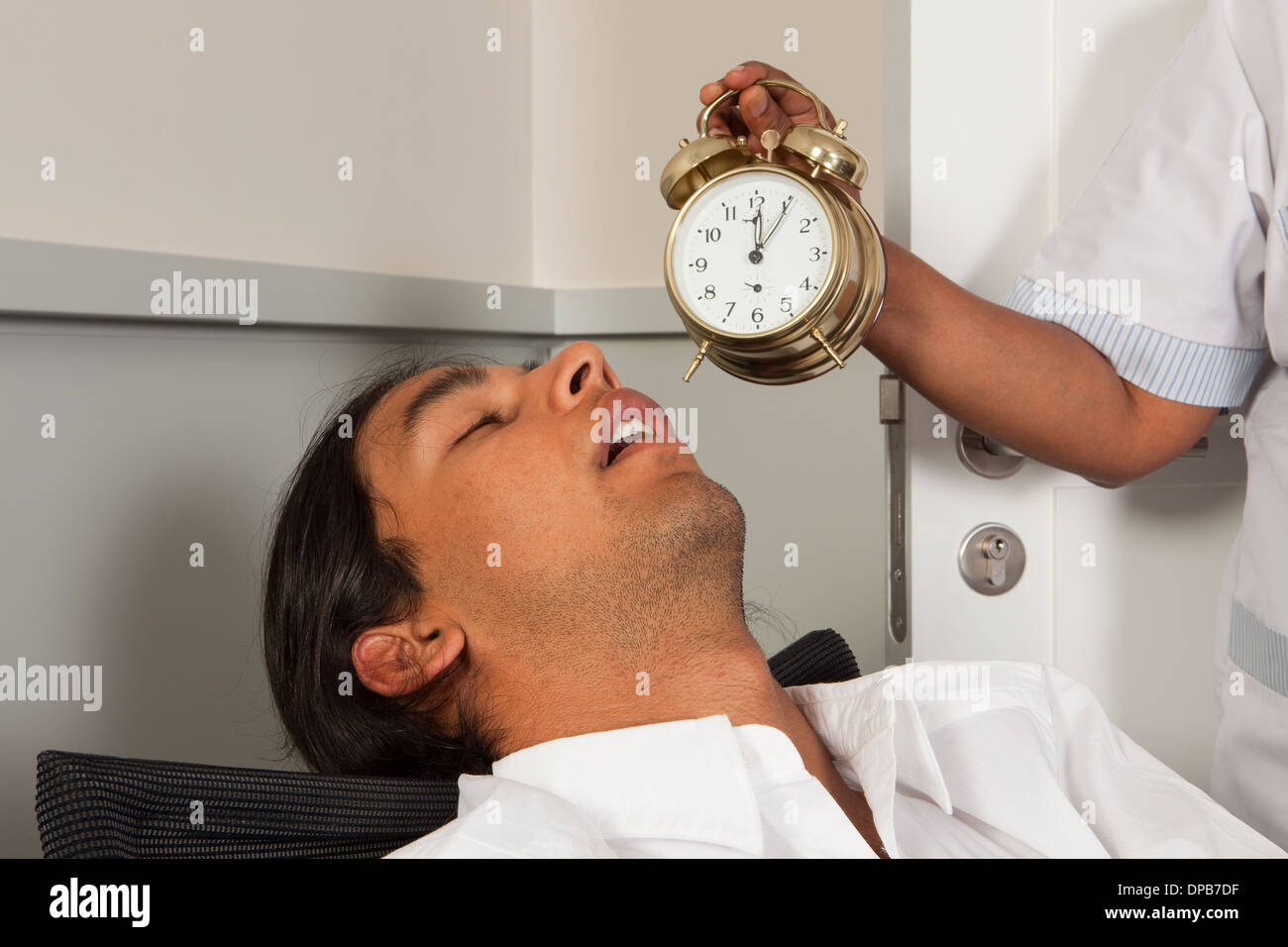 Cleaning woman holding an alarm clock next to a sleeping office worker ...