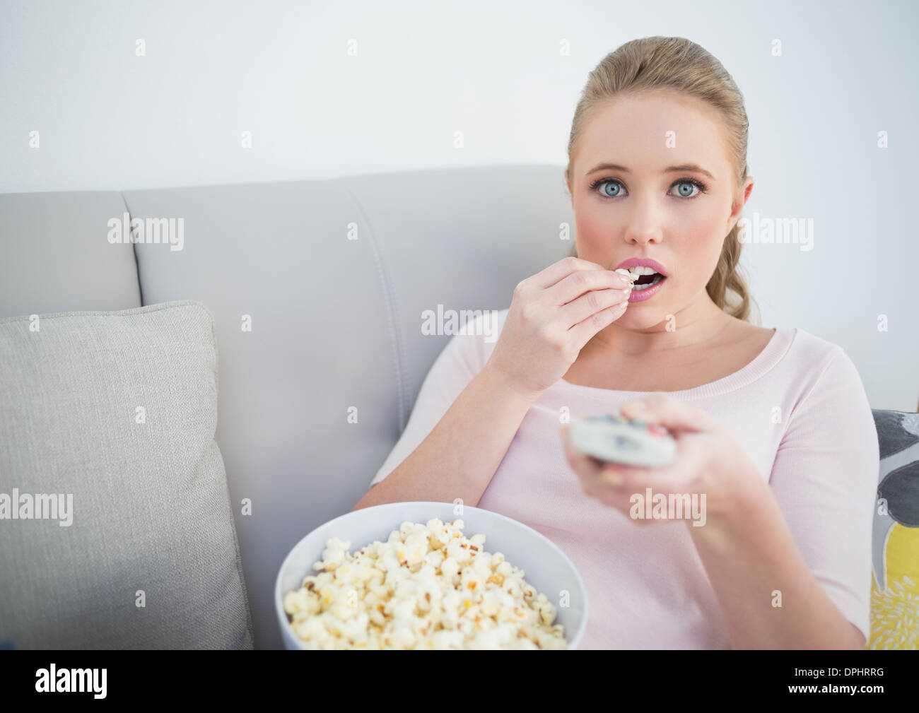 Casual astonished blonde holding remote and eating popcorn Stock Photo ...