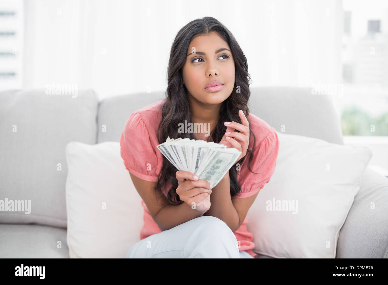Thoughtful cute brunette sitting on couch holding money Stock Photo - Alamy