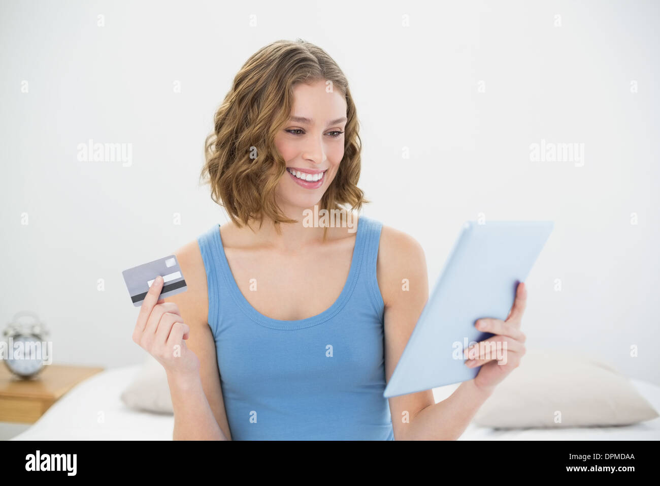 Gorgeous woman holding her tablet and debit card sitting on her bed ...