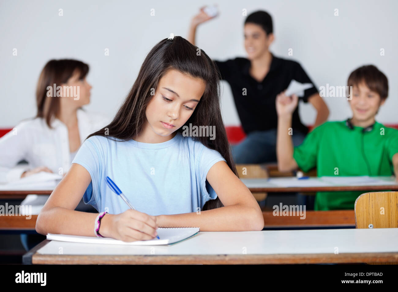 Schoolgirl Writing At Desk While Classmates Playing Stock Photo - Alamy