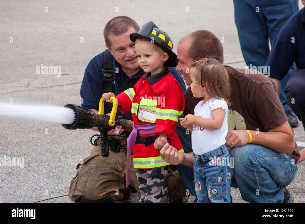 A young boy using a fire hose to spray water with a firefighter and his ...