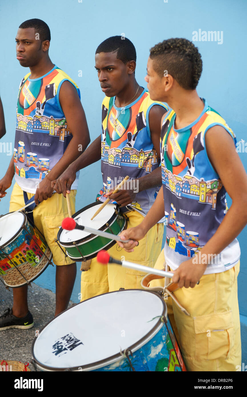 Three young Brazilian men standing drumming against a blue wall in ...