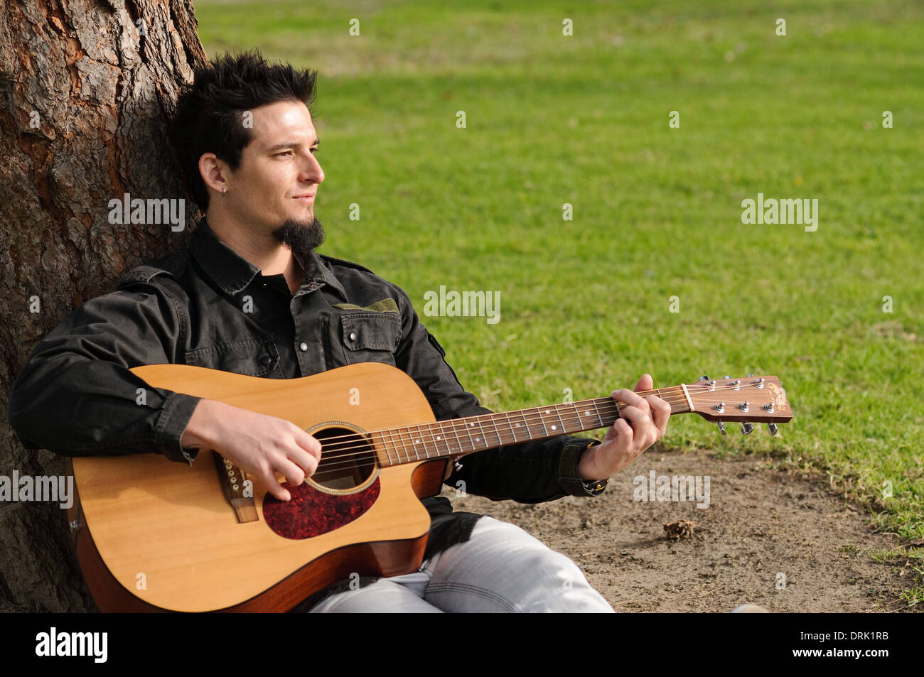 A young man sitting against a tree at a park playing guitar Stock Photo ...