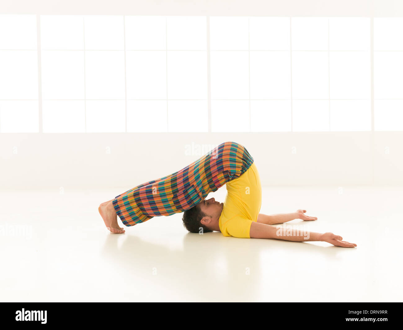 Colorful dressed male repeating Halasana yoga exercises in a white room ...