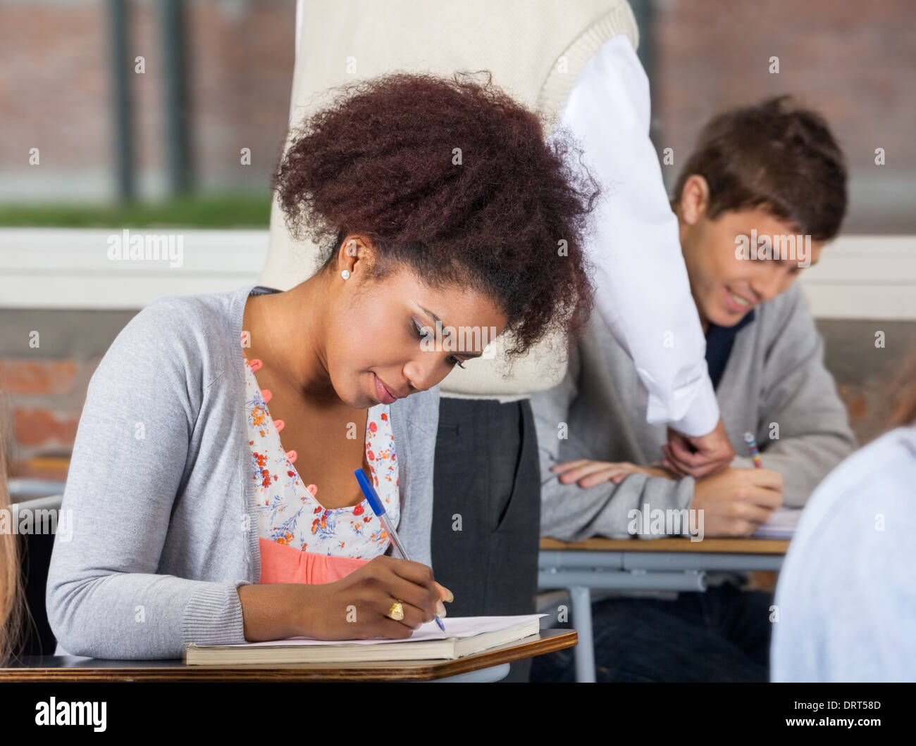Student Writing Exam While Teacher Explaining Classmate In Backg Stock ...