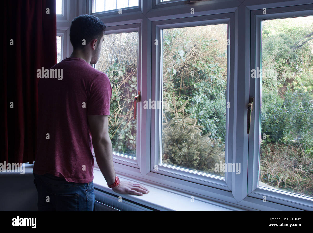 Young man indoors looking out through a window, over shoulder view ...