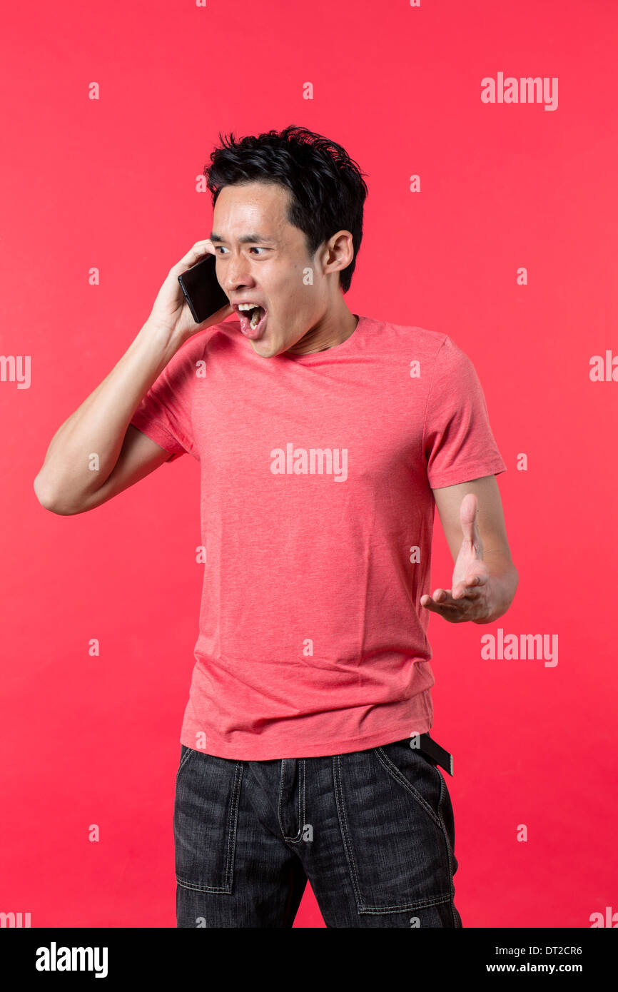 Angry Asian man talking on cell phone. Standing against red background ...