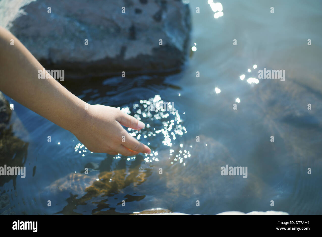 A young girl leaning forward to put her hand into clear shallow lake ...