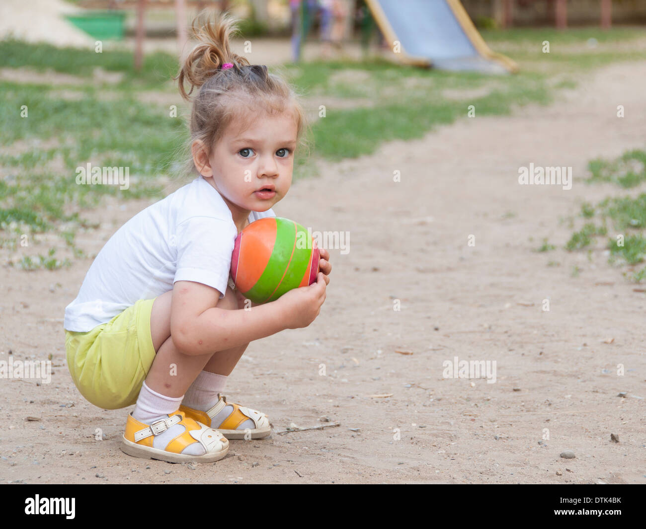 Little girl sitting on his haunches with a ball on the Playground ...