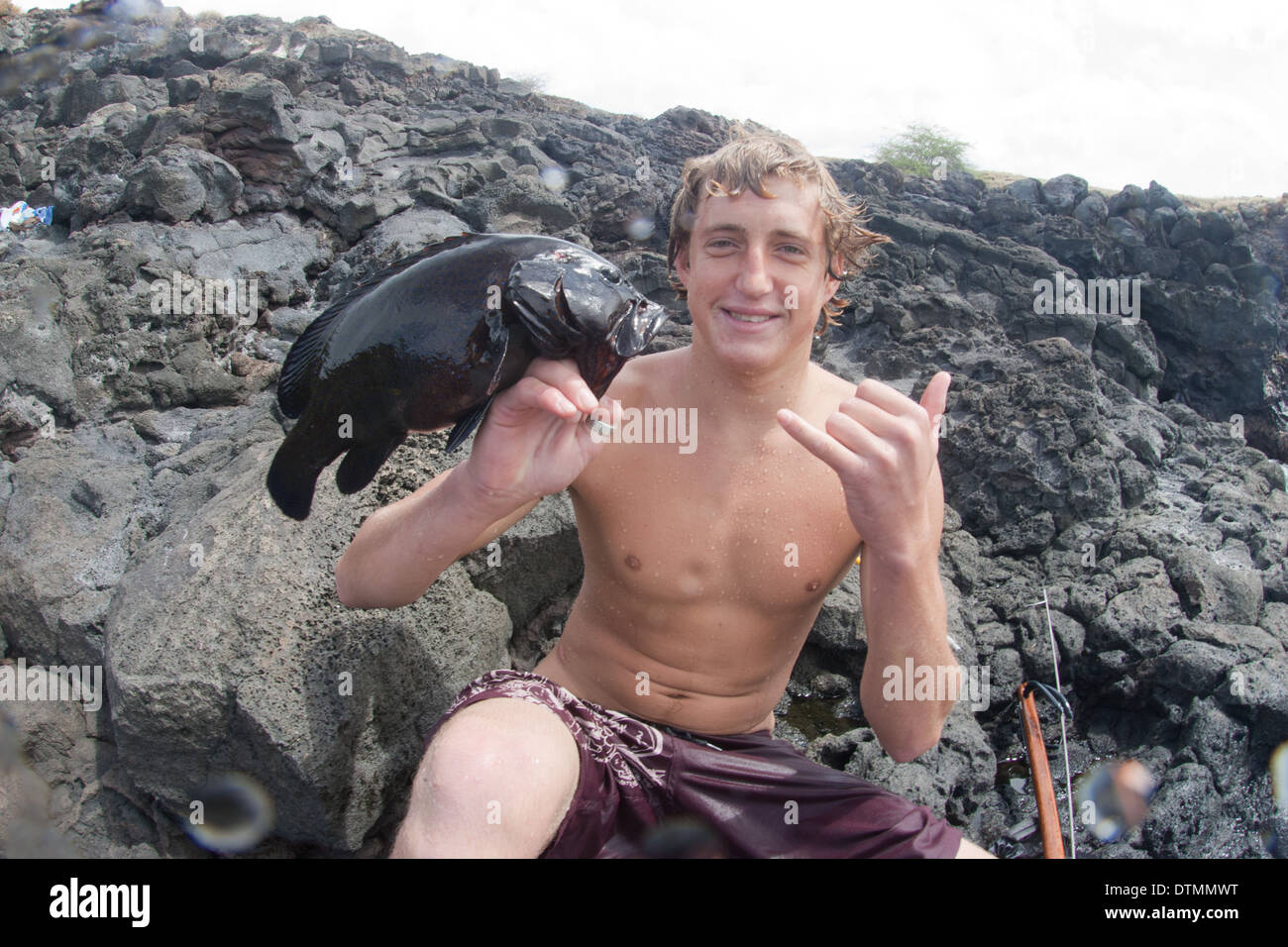 man with a hang loose hand gesture holding a black fish while he sits ...