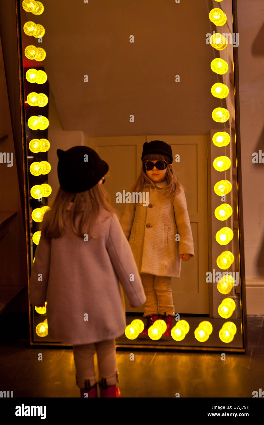 A little girl in sunglasses looking at her reflection in a mirror ...