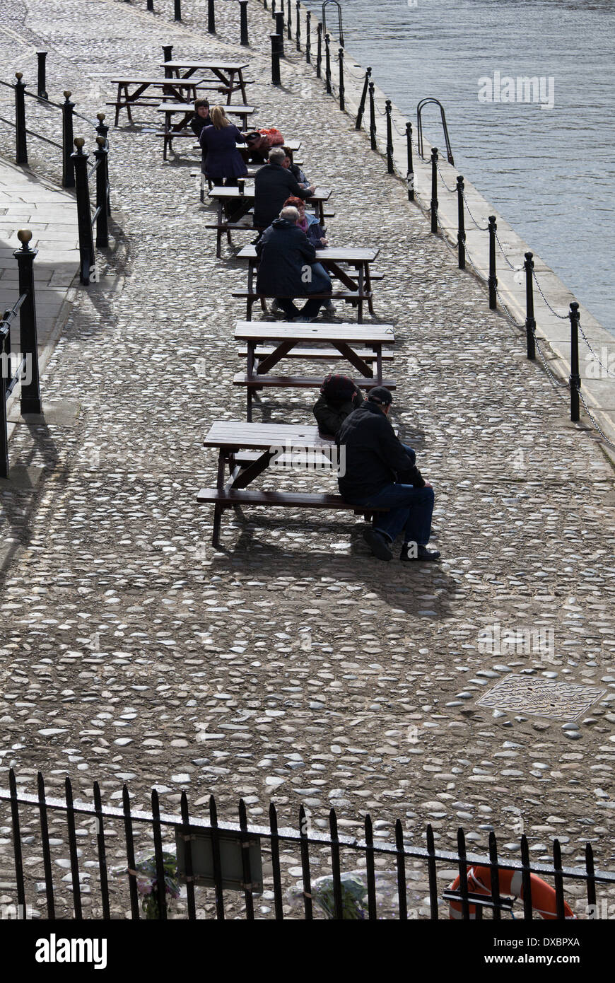 Riverside bar wooden tables and chairs with seated patrons on cobbled ...