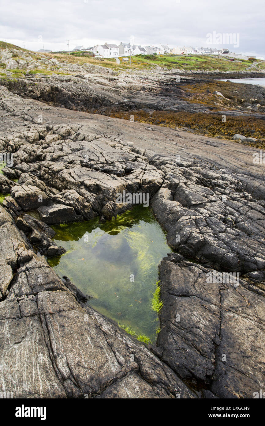 Heart-shaped rock pool in metamorphic rock with village of Portnahaven ...