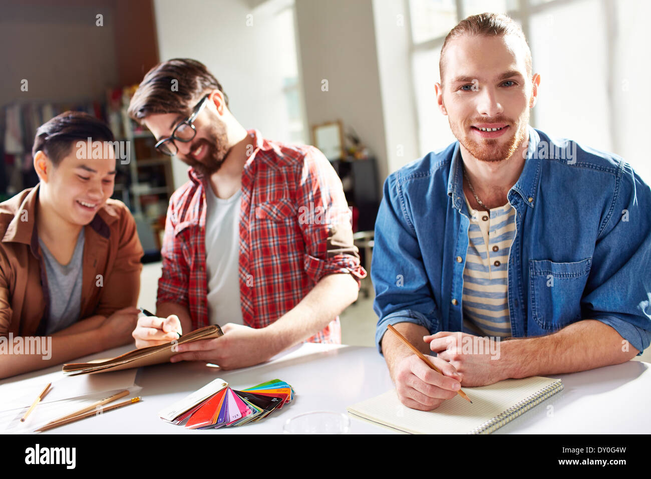 Smiling guy with pencil and notepad looking at camera with his friends ...