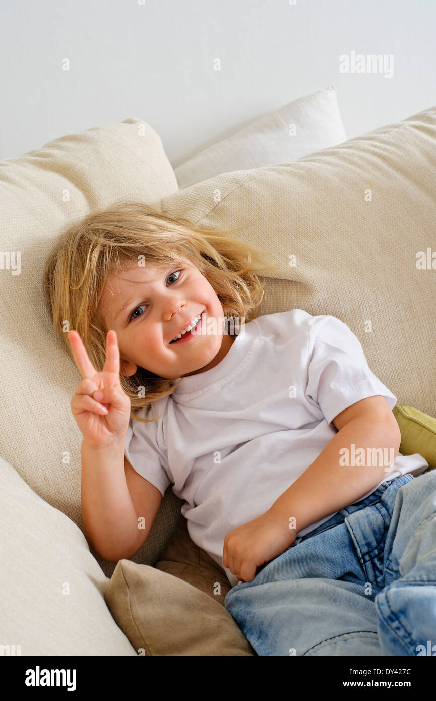 Young boy sitting on sofa looking at camera. He's giving the V sign ...