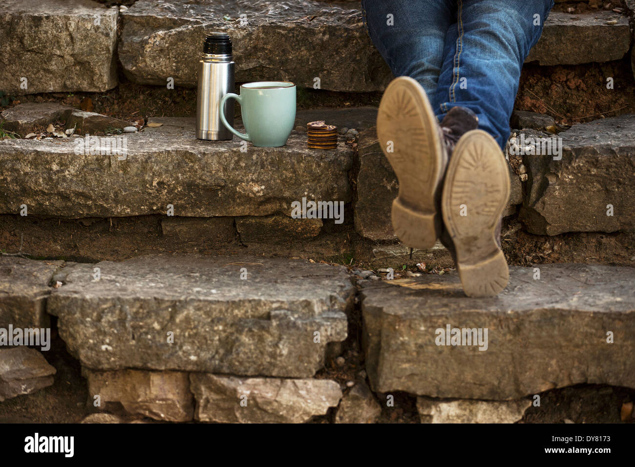 Feet of young man sitting on steps with cup of coffee and thermos flask ...