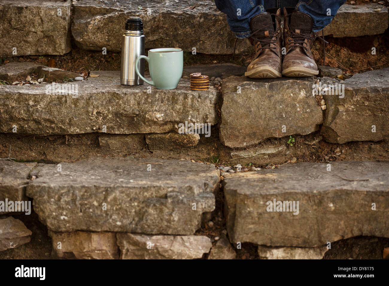 Feet of young man sitting on steps with cup of coffee and thermos flask ...
