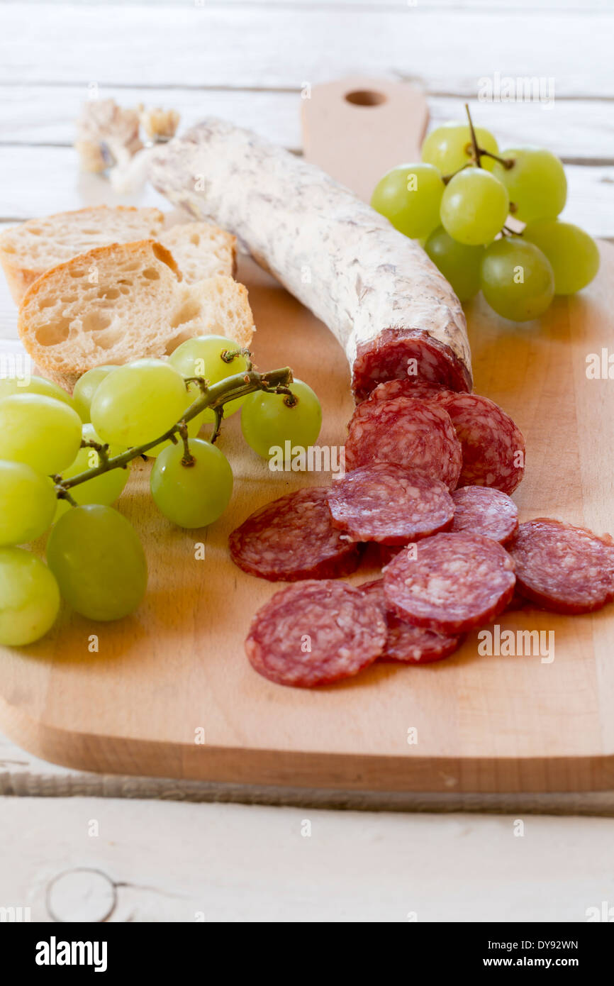 Chopped salami, green grapes, tomatoes and baguette on chopping board ...