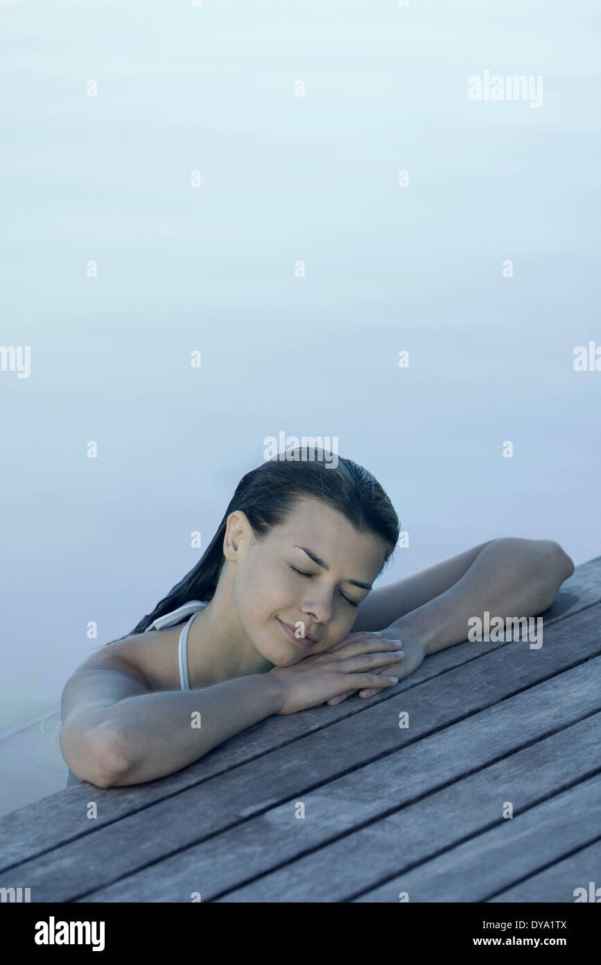 Woman in pool leaning against deck, resting head on arms, eyes closed ...