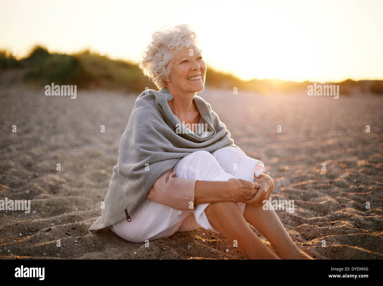 Happy retired woman wearing shawl sitting relaxed on sand at the beach ...