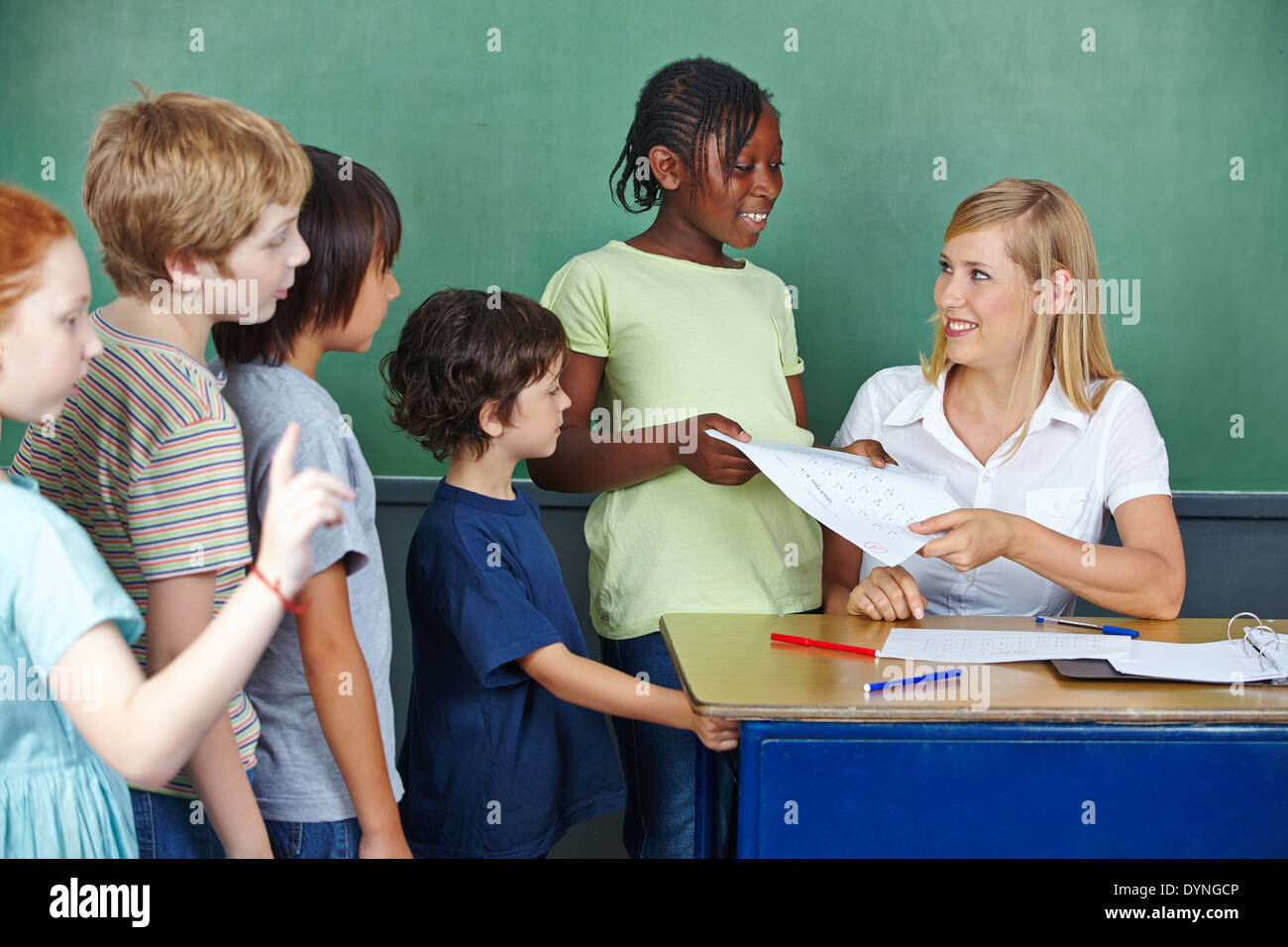 Elementary school students in a row at desk of teacher to get their ...