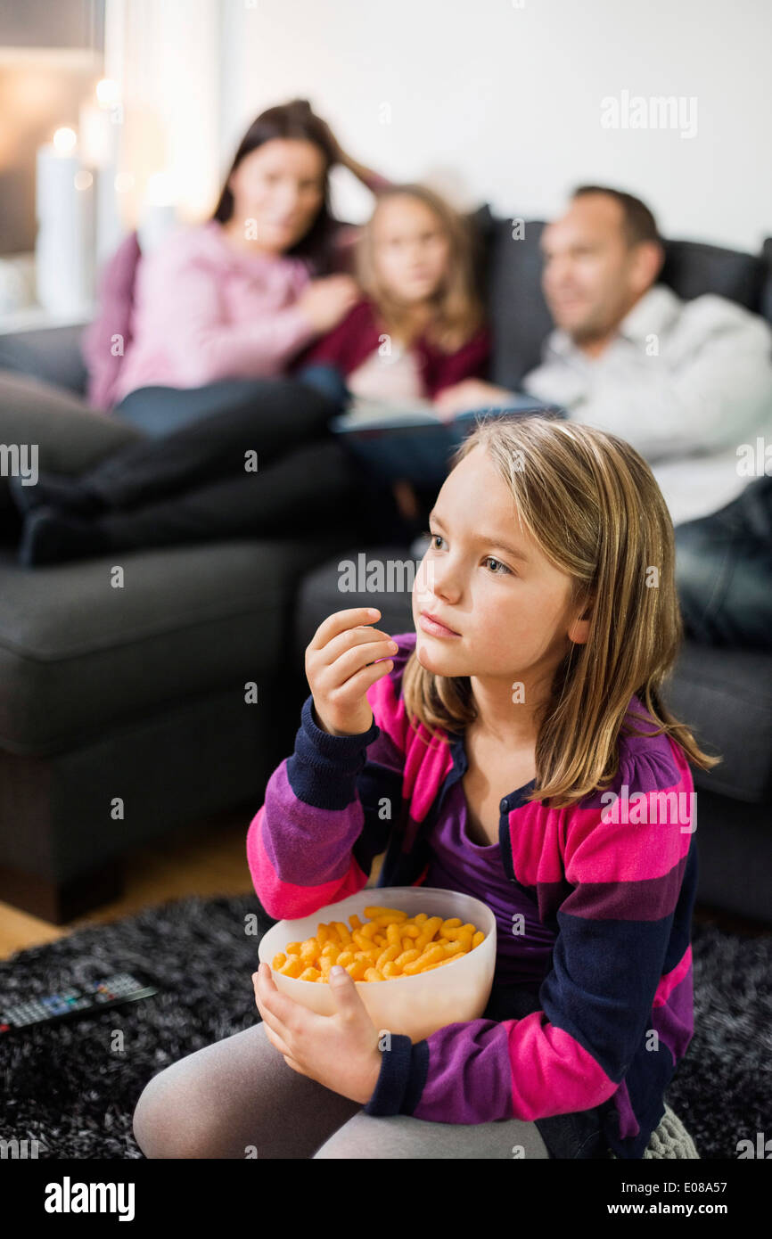 Girl having snacks while watching TV on floor with family in background ...