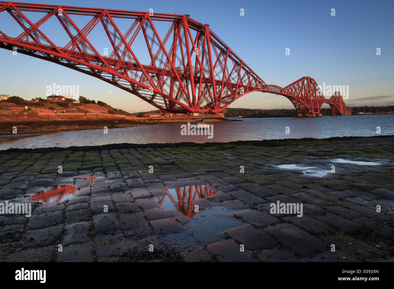 The Forth Bridge at sunset, reflected in puddles on the slipway at ...