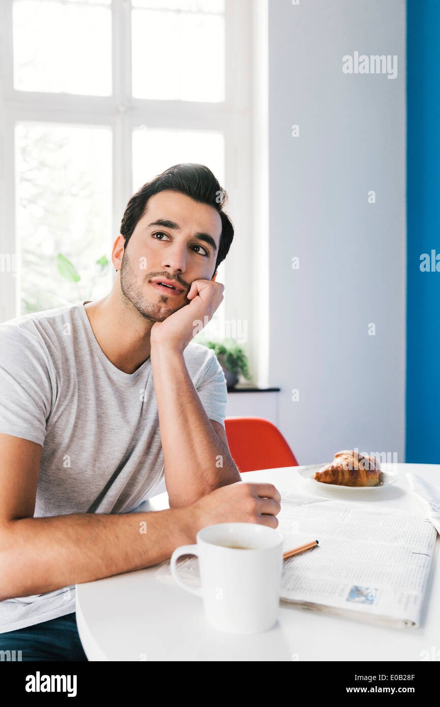 Portrait of pondering young man sitting at breakfast table with ...
