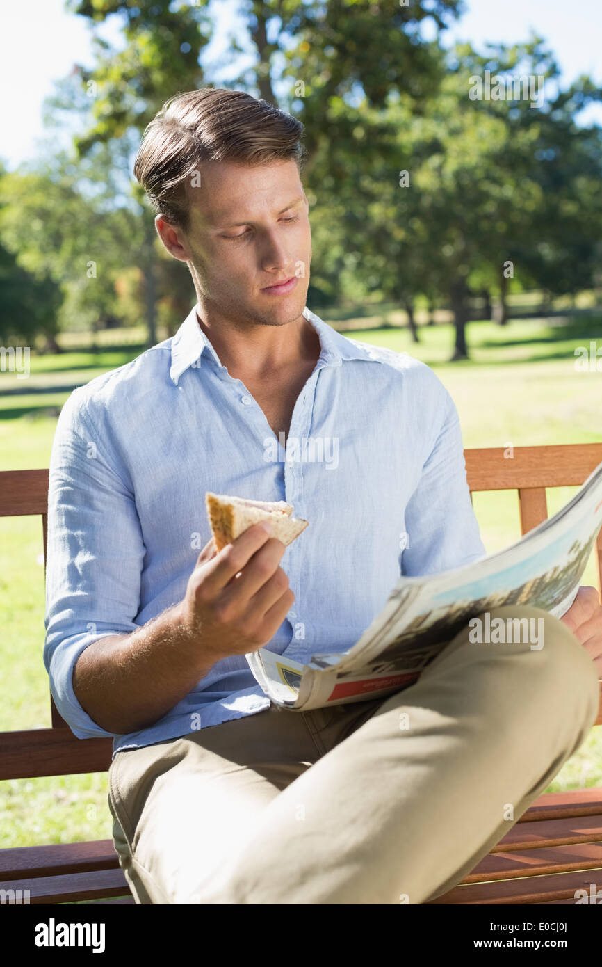 Handsome man sitting on park bench eating sandwich and reading paper ...
